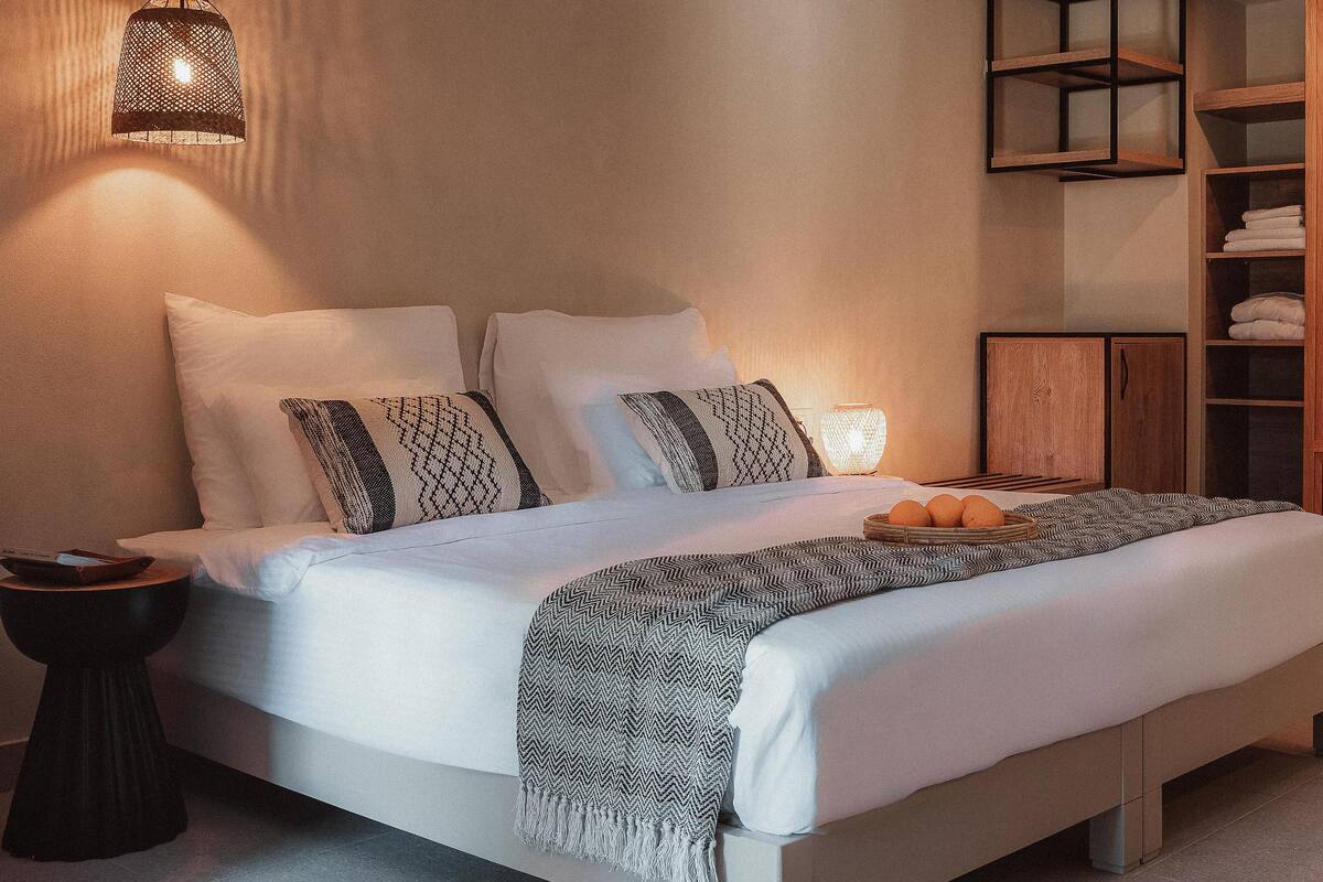 Uma cama bem arrumada com lençóis brancos e almofadas estampadas, uma mesa de cabeceira com abajur e uma estante de madeira com toalhas dobradas.