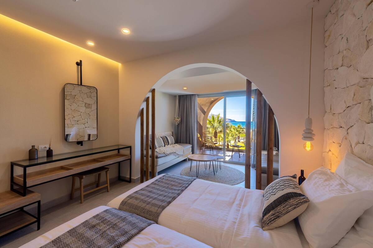 Quarto moderno com uma cama grande, uma escrivaninha e um sofá. Há vista para o exterior através de uma porta de vidro.