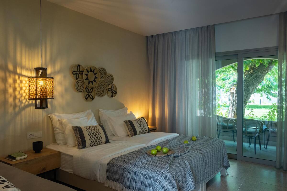 Quarto com cama, mesa de cabeceira e vista do exterior através de uma porta de vidro.