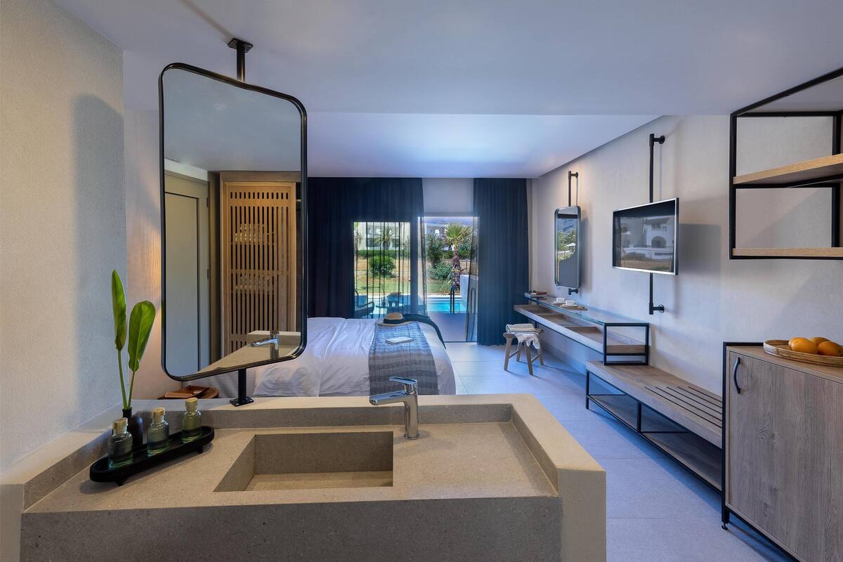 Quarto de hotel moderno com uma cama grande, toucador com espelho, pia e escrivaninha. Vê-se uma piscina e vegetação lá fora.