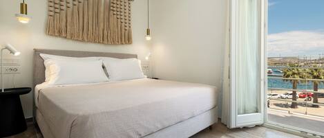 Quarto com cama, luminárias de cabeceira, um painel de macramé na parede e vista para o mar por uma porta aberta.