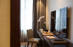 Quarto de hotel com um espelho grande, uma escrivaninha com um laptop, uma cadeira e um vaso com flores.