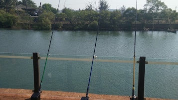 Uma área de pesca com três varas de pesca fixadas em suportes, um primeiro plano gramado e um corpo d'água calmo com uma linha de costa ao longe.
