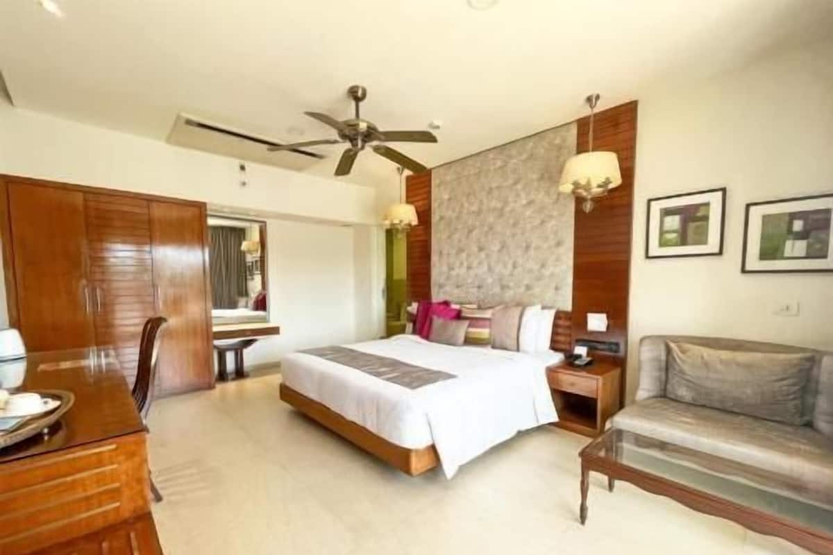 Quarto de hotel com cama, escrivaninha, sofá, ventoinha no teto e luminária na parede.