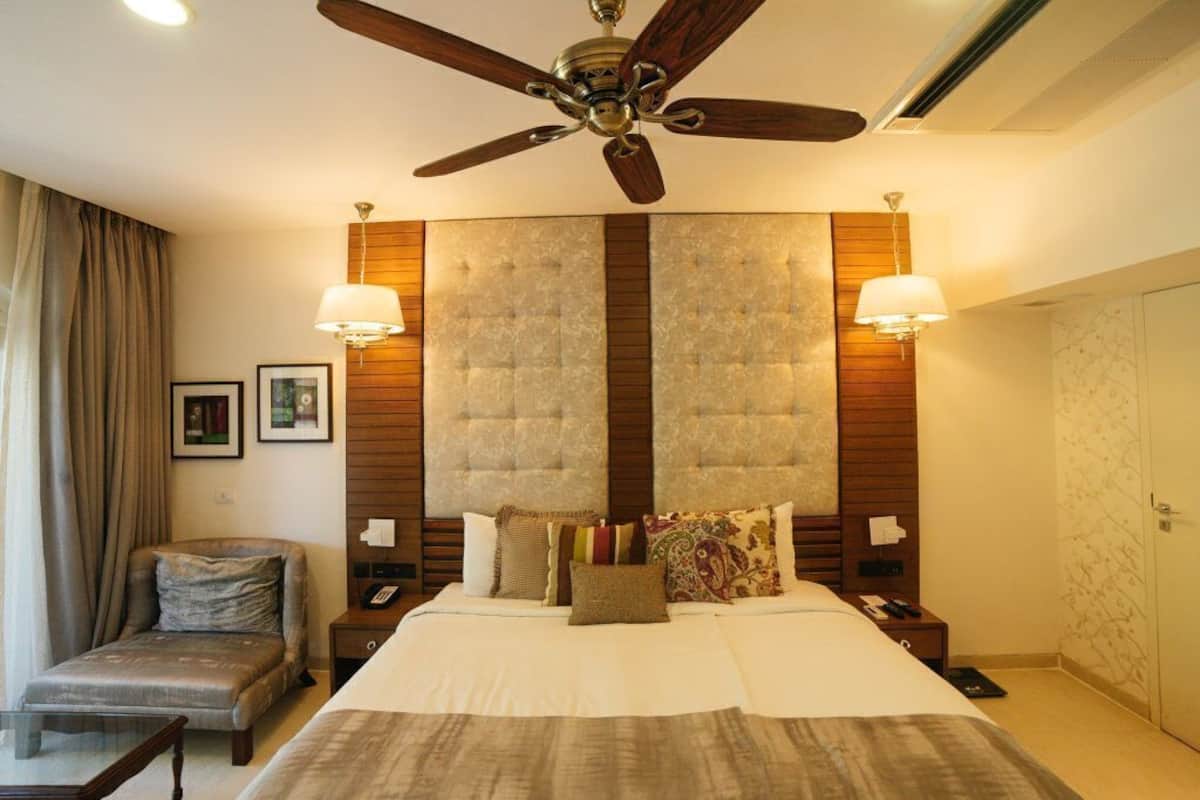 Quarto de hotel com uma cama grande, um ventilador de teto, duas luminárias de cabeceira, uma mesinha de cabeceira, uma cadeira e um guarda-roupa.