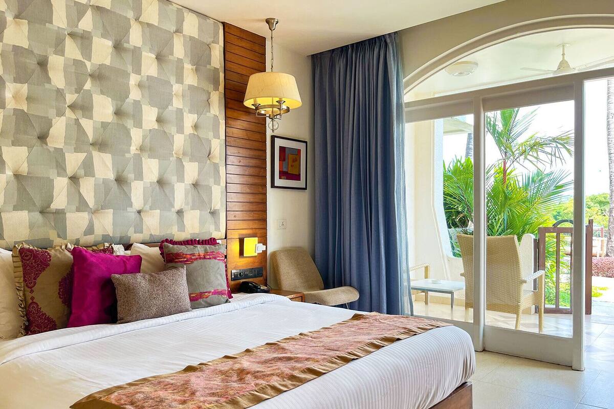 Quarto de hotel com uma cama grande, cabeceira de madeira, ventilador de teto, vista para o exterior e uma varanda com cadeiras.