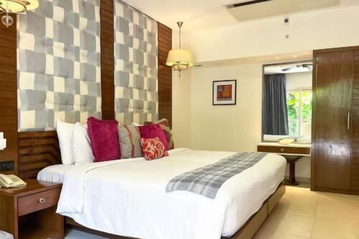Quarto de hotel com uma cama grande, mesa de cabeceira com luminária, cabeceira de madeira, janela com cortinas e uma porta.