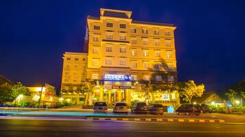Hotel de vários andares com uma placa visível, vários carros estacionados e uma entrada bem iluminada.