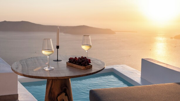 Mesa redonda de madeira com duas taças de vinho e uma vela, com vista para uma piscina e um pôr do sol.