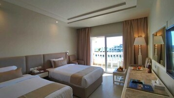 Quarto de hotel com duas camas, uma escrivaninha com luminária, televisão e varanda com vista.