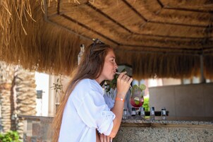 Uma pessoa num bar com uma bebida, um telhado de palha e uma parede de pedra.