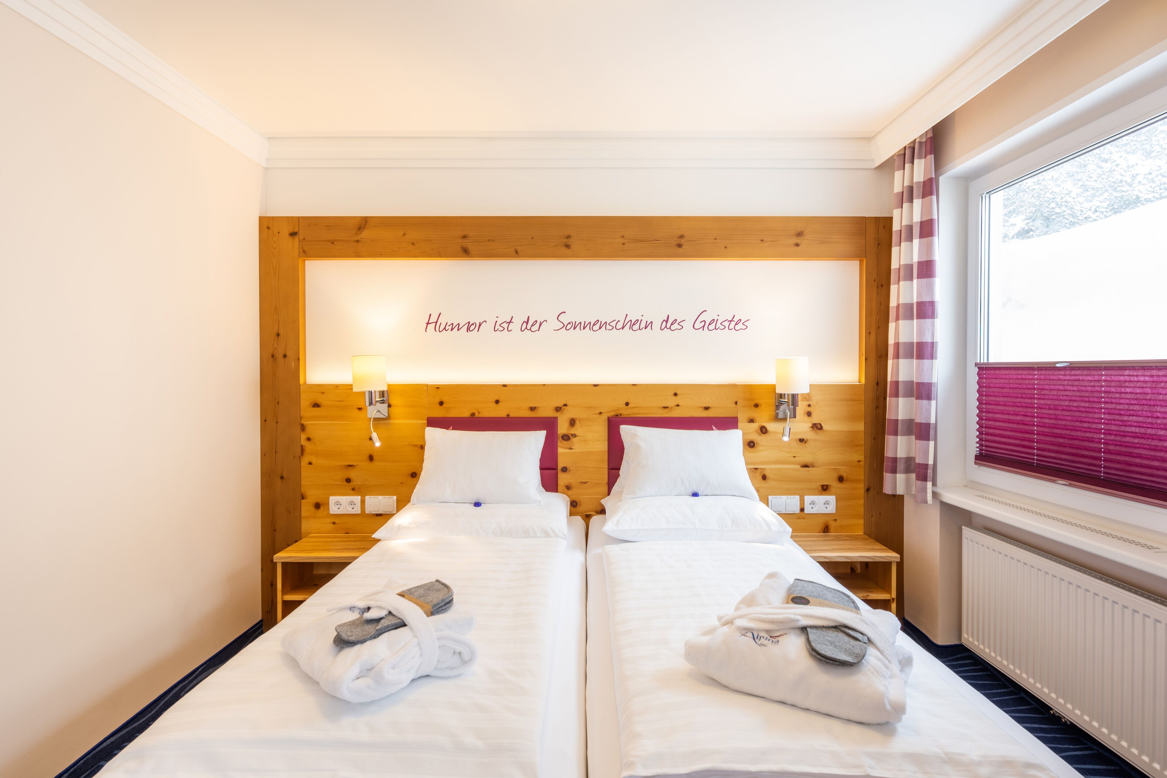 Quarto de hotel com duas camas, cabeceira de madeira, roupa de cama branca, toalhas, abajures e uma janela com persianas.