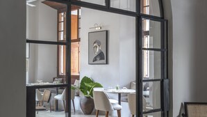 Um interior moderno com uma grande janela em arco, uma área de jantar com cadeiras e uma mesa, e uma grande planta em vaso.