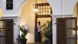 Entrada de hotel com placa, venezianas de madeira e plantas em vasos.