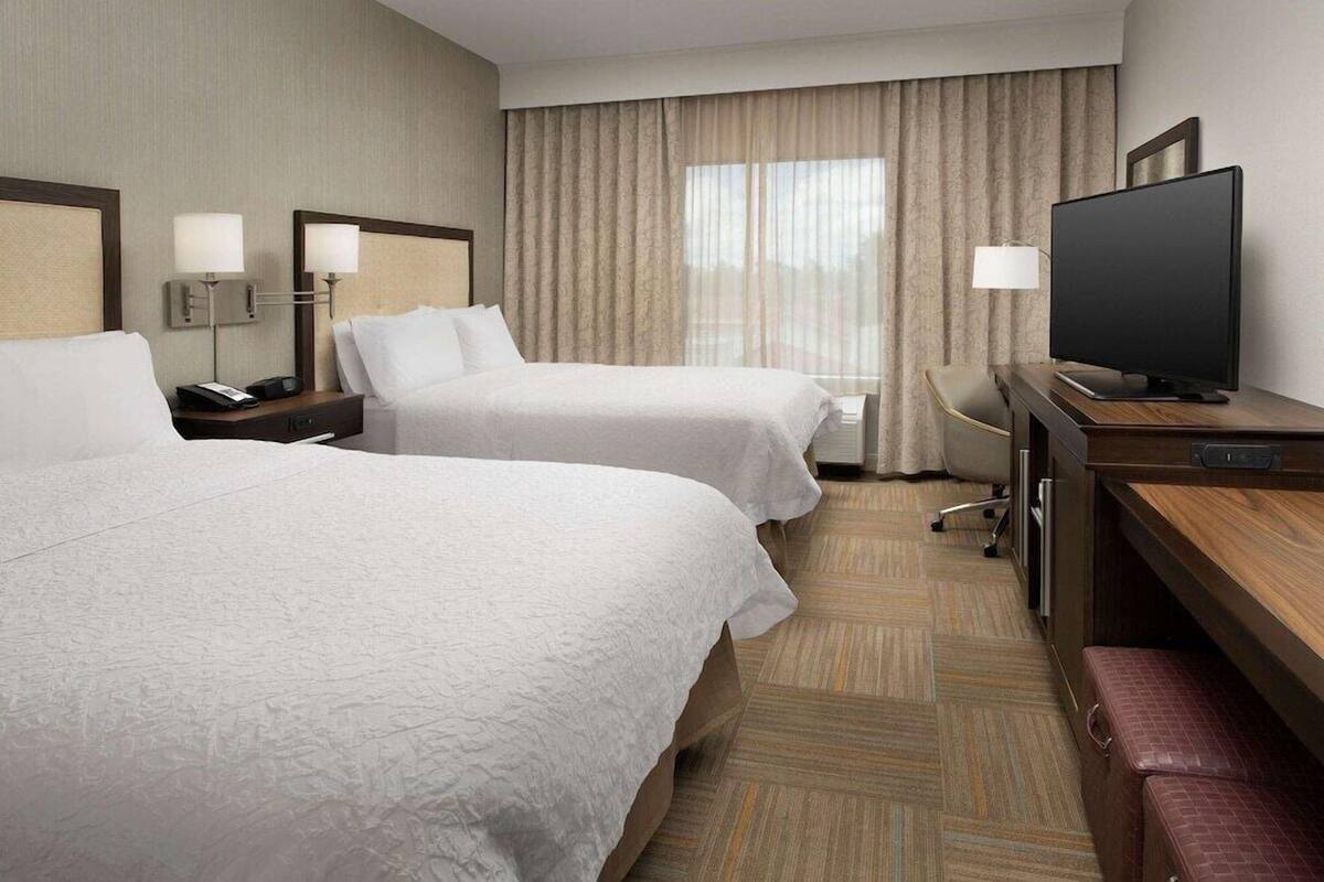 Quarto de hotel com duas camas, uma escrivaninha com televisão, uma cadeira e uma luminária.