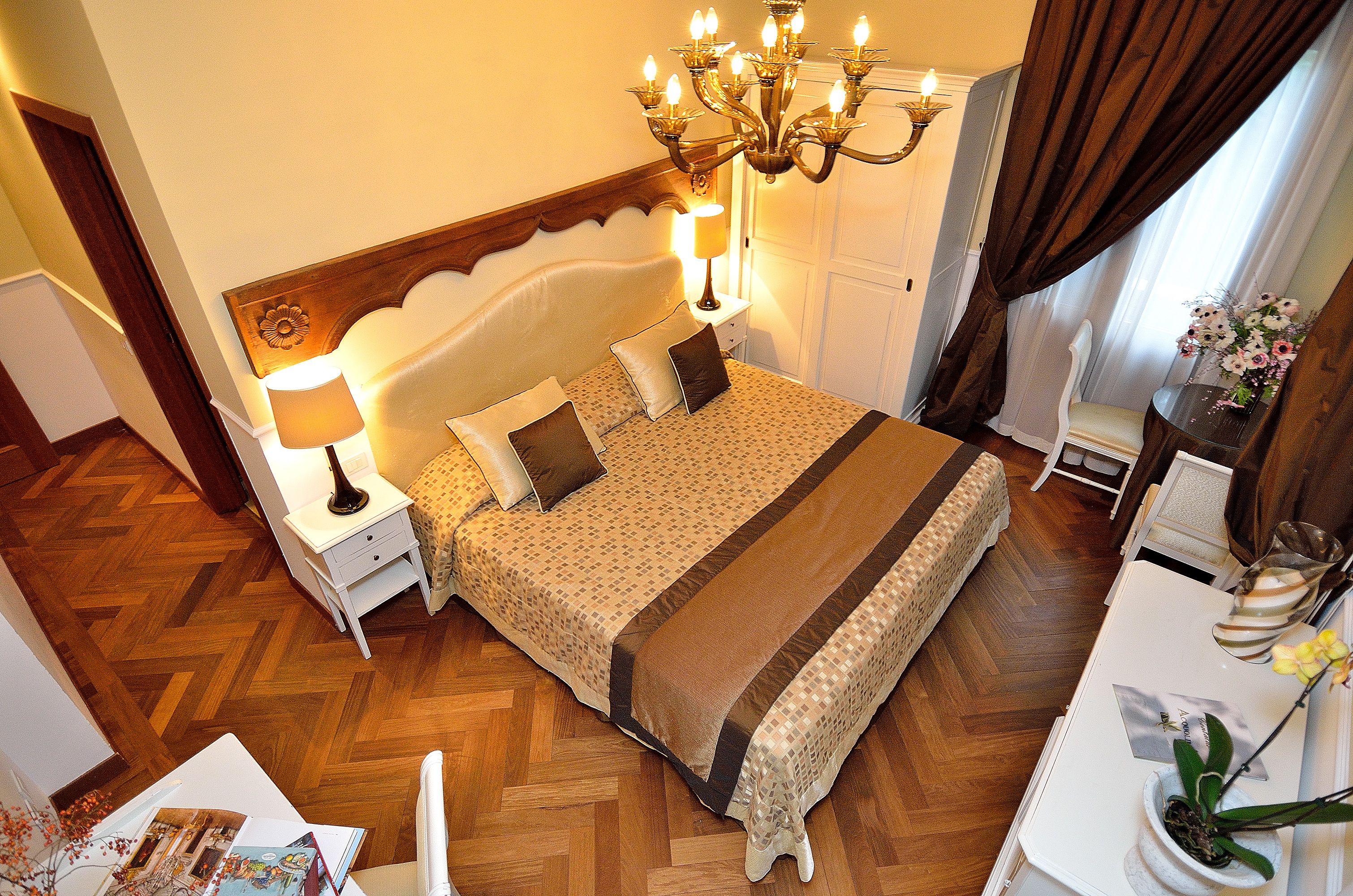Quarto de hotel com uma cama grande, um lustre, uma mesa de cabeceira, uma cadeira e uma pequena mesa com uma planta.