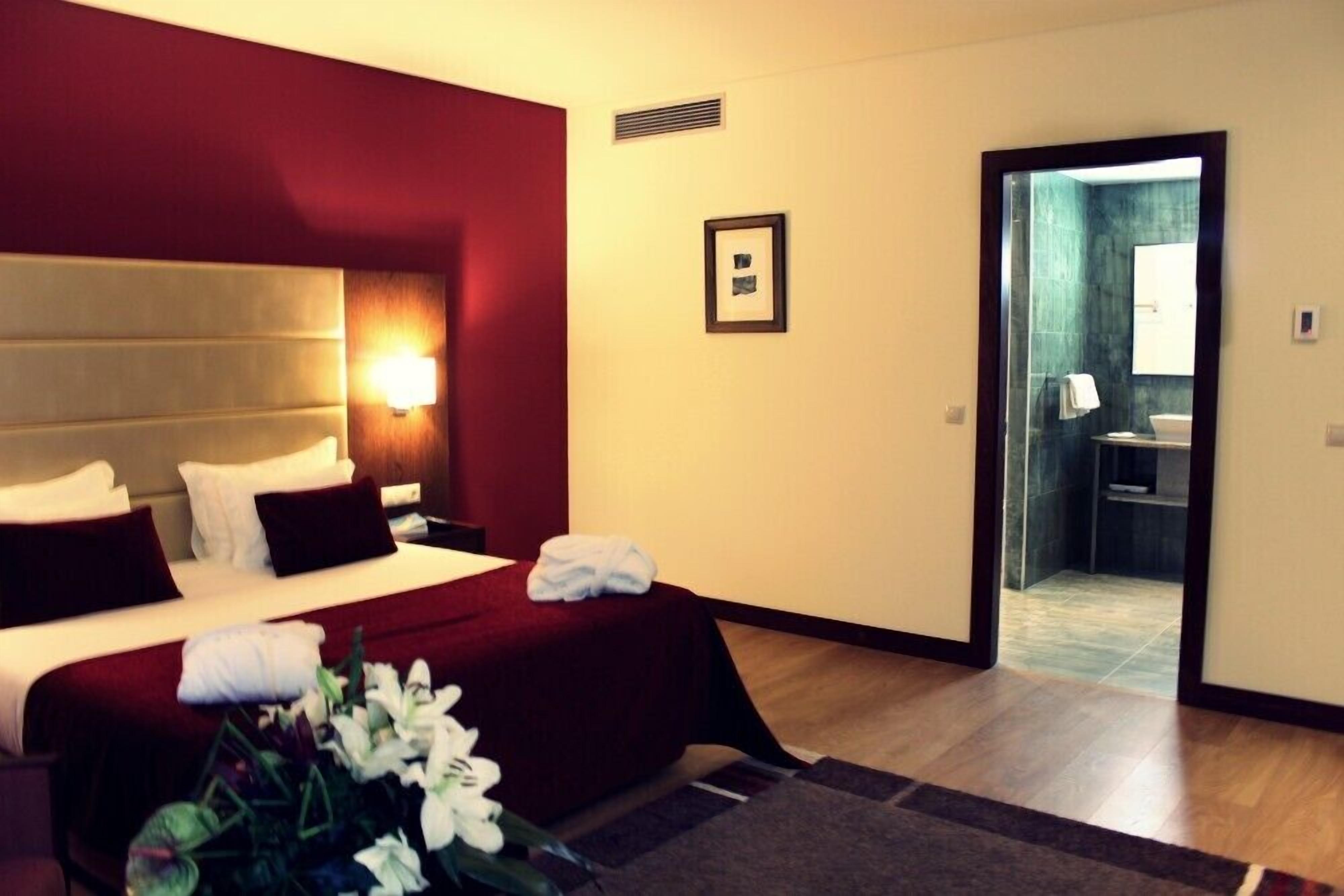 Quarto de hotel com uma cama grande, mesa de cabeceira, luminária na parede, quadro emoldurado e banheiro visível por uma porta aberta.