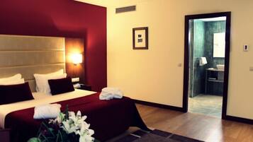 Quarto de hotel com uma cama grande, mesa de cabeceira, luminária na parede, quadro emoldurado e banheiro visível por uma porta aberta.