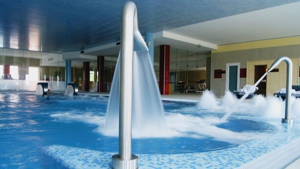 Piscina interior moderna com um recurso aquático central, paredes de vidro transparente e chão revestido com azulejos.