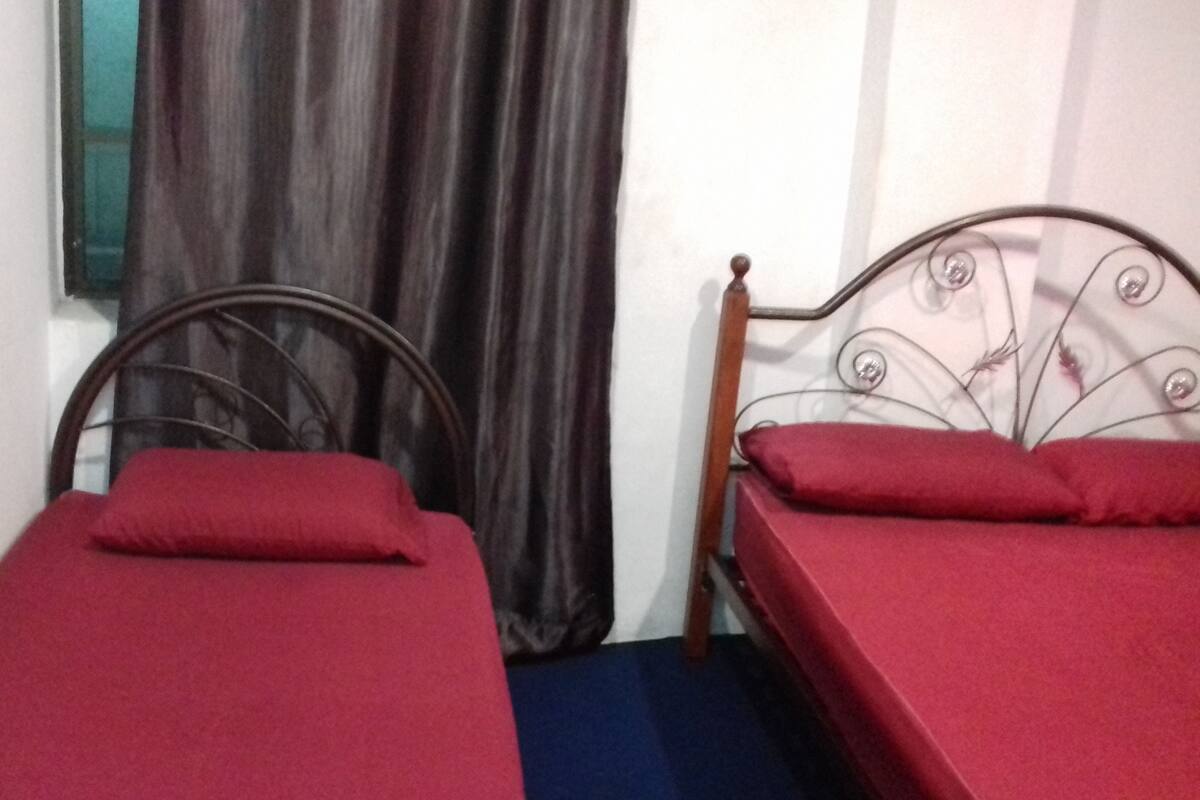 Quarto pequeno com duas camas de solteiro, cada uma com colchão e almofadas vermelhas. As camas têm estrutura metálica com detalhes decorativos. Uma cadeira de madeira está encostada à parede, e uma cortina está parcialmente aberta.