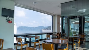 Um restaurante com mesas e cadeiras de madeira, janelas amplas com vista para o mar e colinas, e uma televisão de tela plana na parede.
