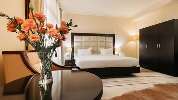 Quarto de hotel com cama, mesas de cabeceira, um vaso com flores, uma mesa redonda e um guarda-roupa.