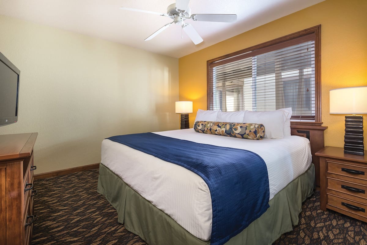 Quarto de hotel com cama, ventoinha no teto, janela com persianas, luminária e cómoda.