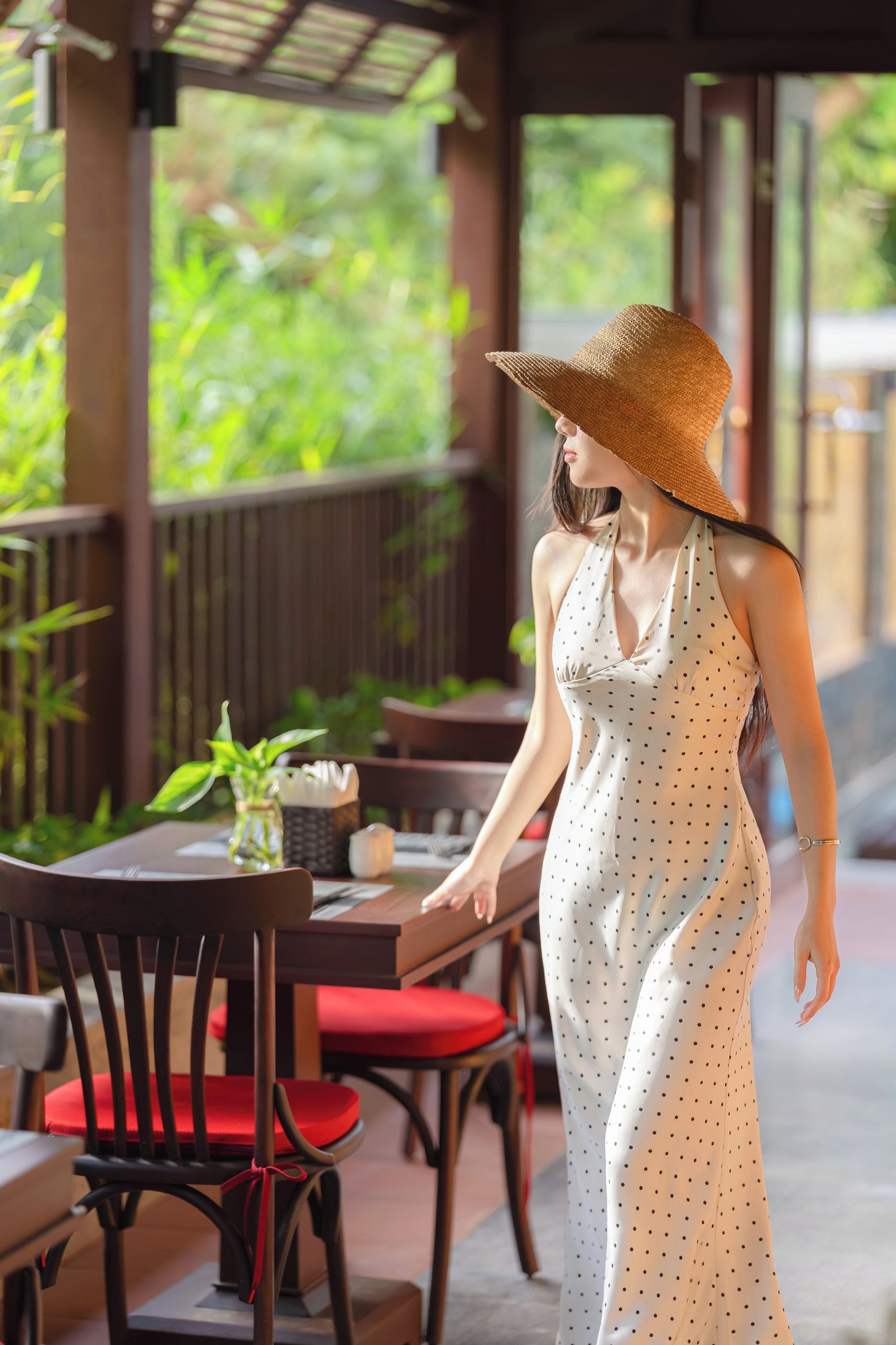 Uma mulher grávida vestida com um vestido de bolinhas, de pé em frente a uma mesa de madeira com um encosto vermelho, em um ambiente externo com vegetação ao fundo.