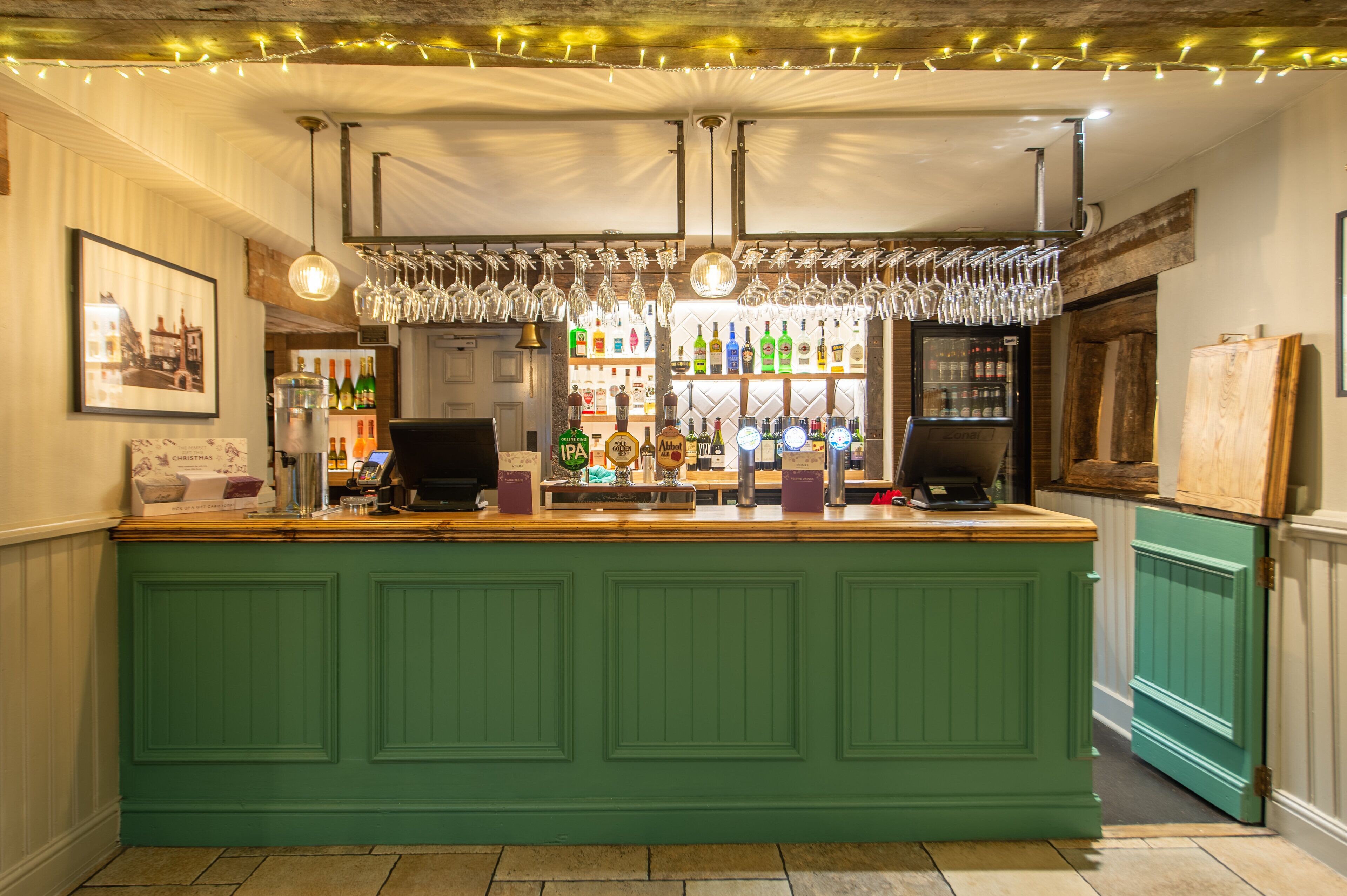 Uma área de bar com balcão verde, copos pendurados e variedade de garrafas.