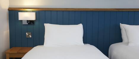 Quarto de hotel com cabeceira azul, duas camas, mesa de cabeceira, luminária acoplada à parede e quadros na parede.