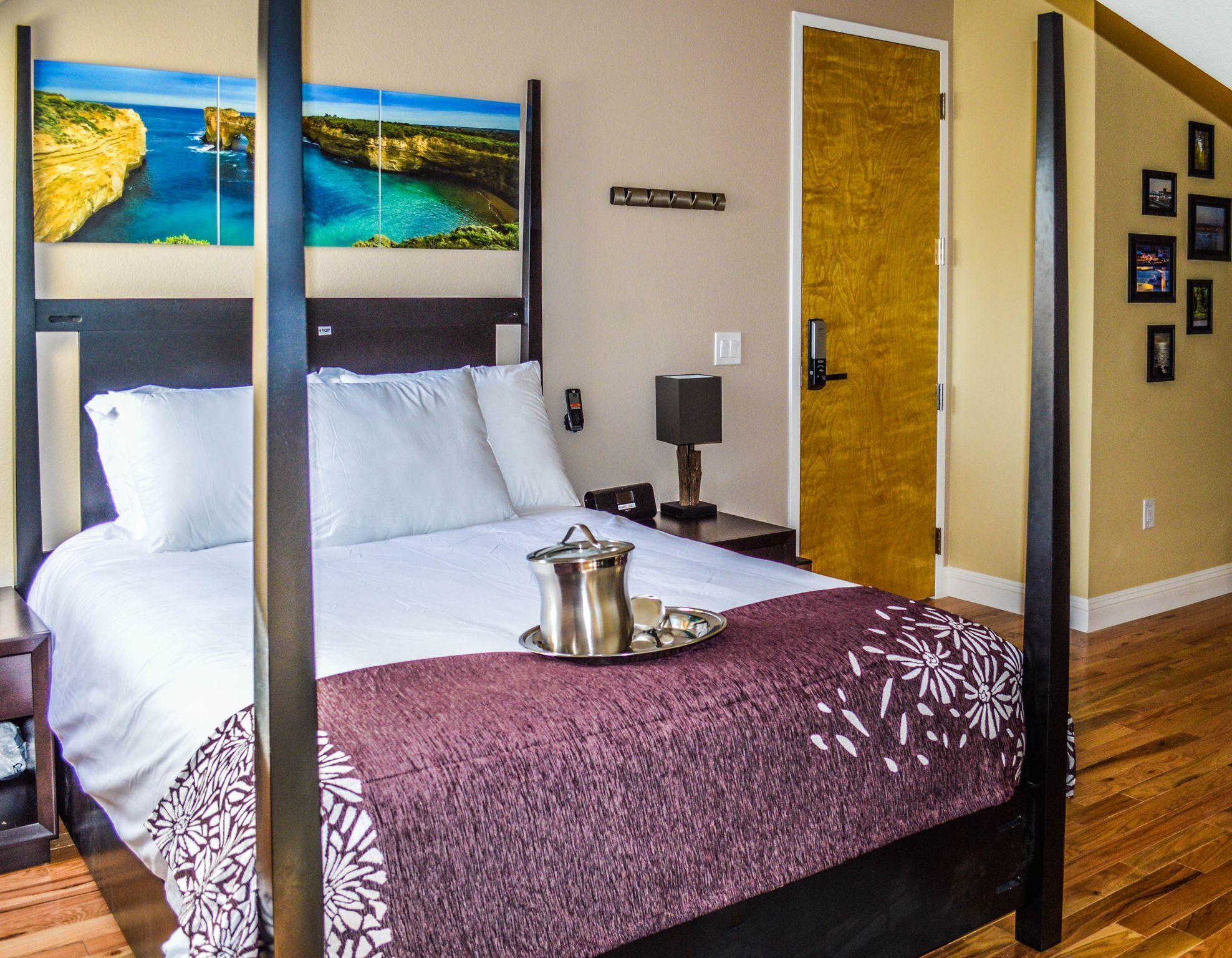 Quarto de hotel com cama, mesa de cabeceira, luminária, bandeja com bule e pintura de uma paisagem costeira.
