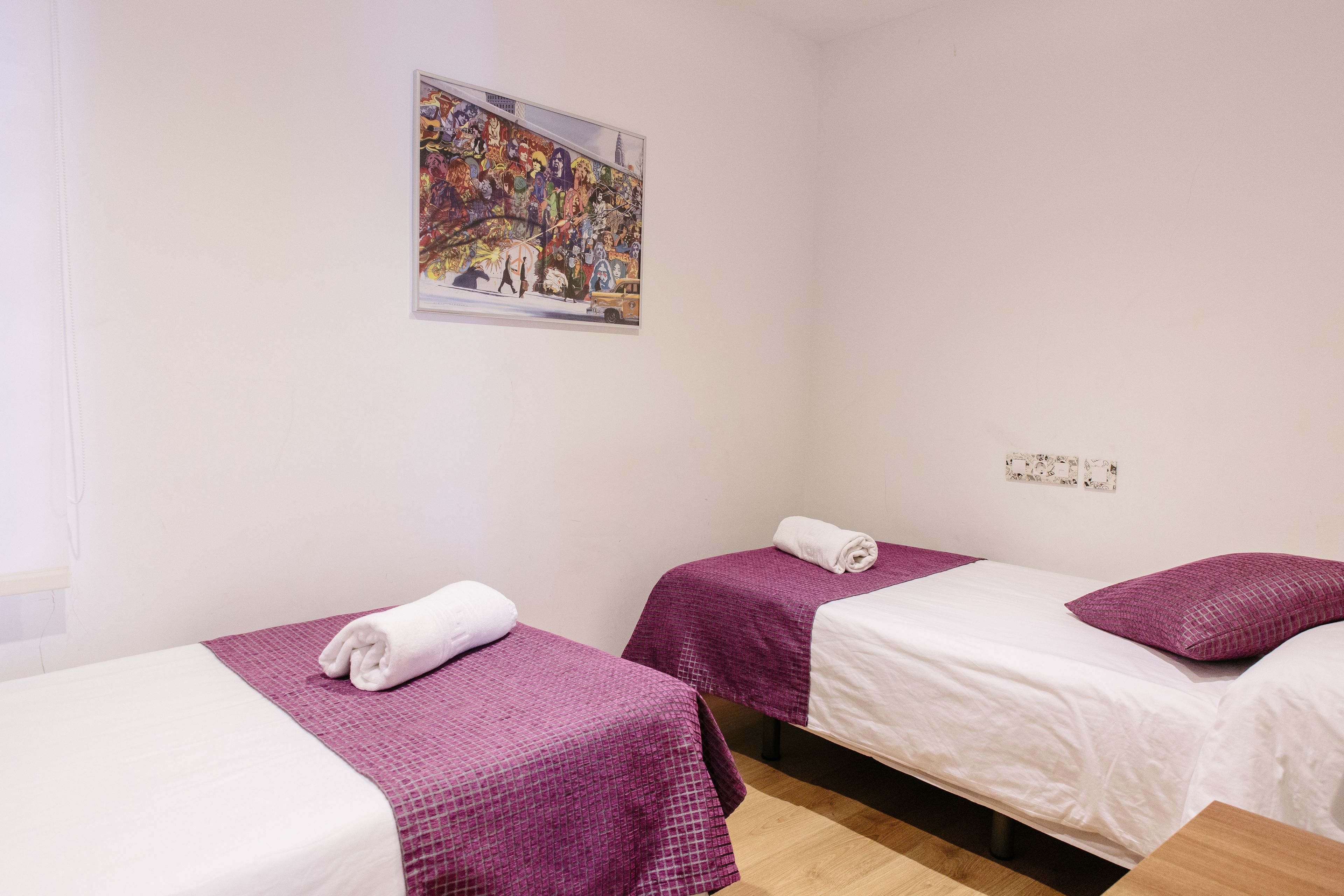 Quarto com duas camas, cada uma com colcha roxa e roupa de cama branca. Há um cartaz colorido na parede acima das camas.