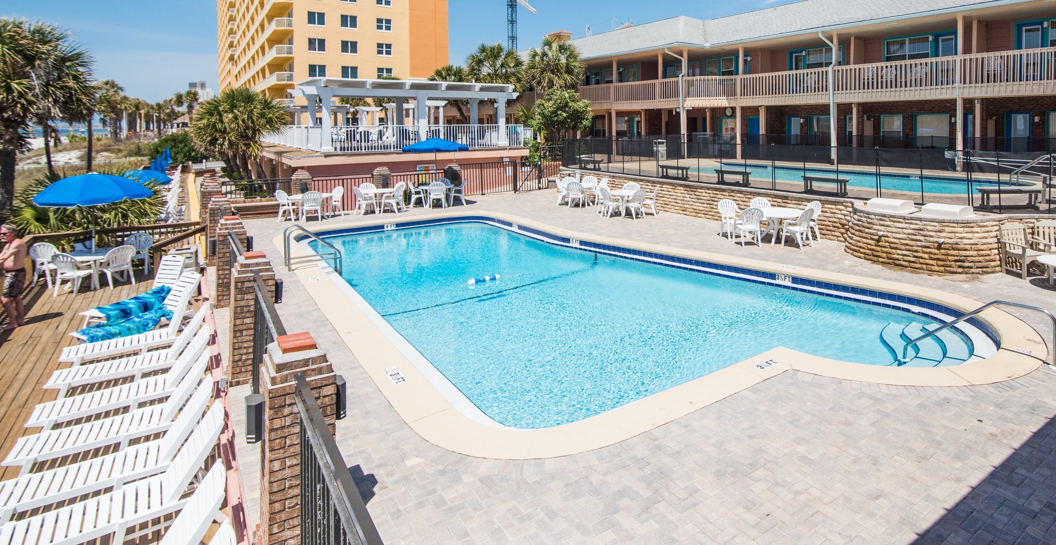 Uma área de piscina com espreguiçadeiras e guarda-sóis, um espaço de estar coberto e um edifício com várias varandas.