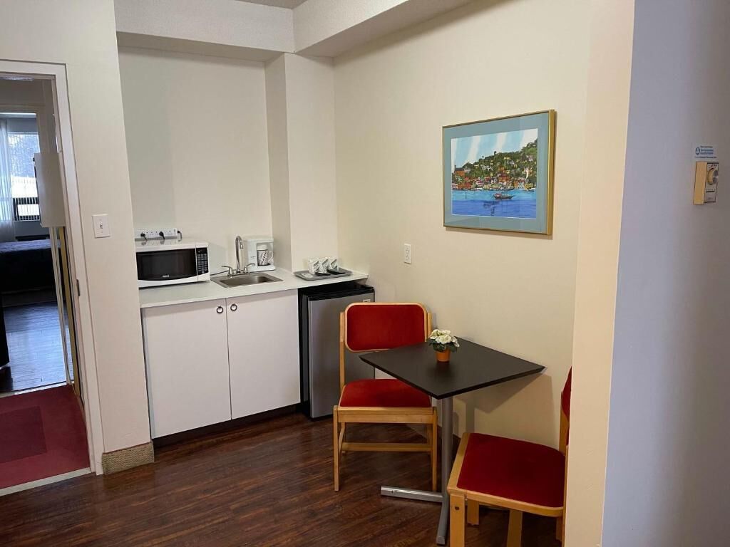 Cozinha compacta com armários brancos, micro-ondas e pia. Possui área de refeições com uma mesa pequena e duas cadeiras. Um quadro com uma paisagem costeira enfeita a parede.