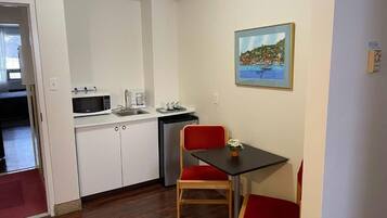 Cozinha compacta com armários brancos, micro-ondas e pia. Possui área de refeições com uma mesa pequena e duas cadeiras. Um quadro com uma paisagem costeira enfeita a parede.