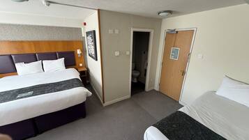 Quarto de hotel com duas camas, uma cabeceira de madeira, uma mesa de cabeceira, uma luminária na parede e uma porta que dá para o banheiro.