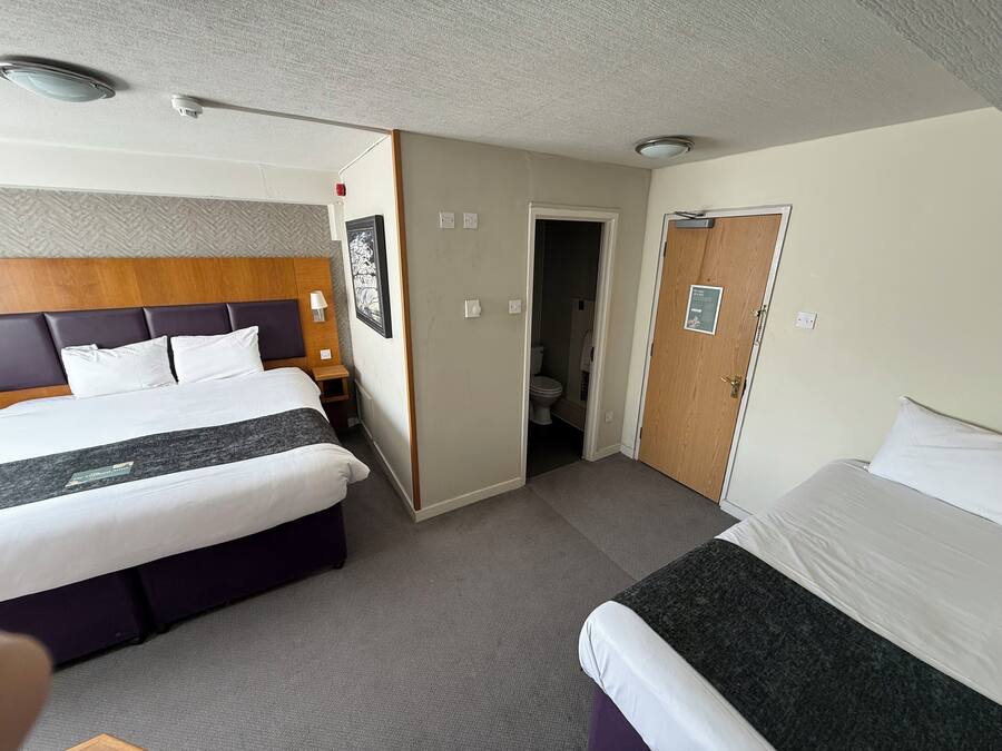 Quarto de hotel com duas camas, uma cabeceira de madeira, uma mesa de cabeceira, uma luminária na parede e uma porta que dá para o banheiro.