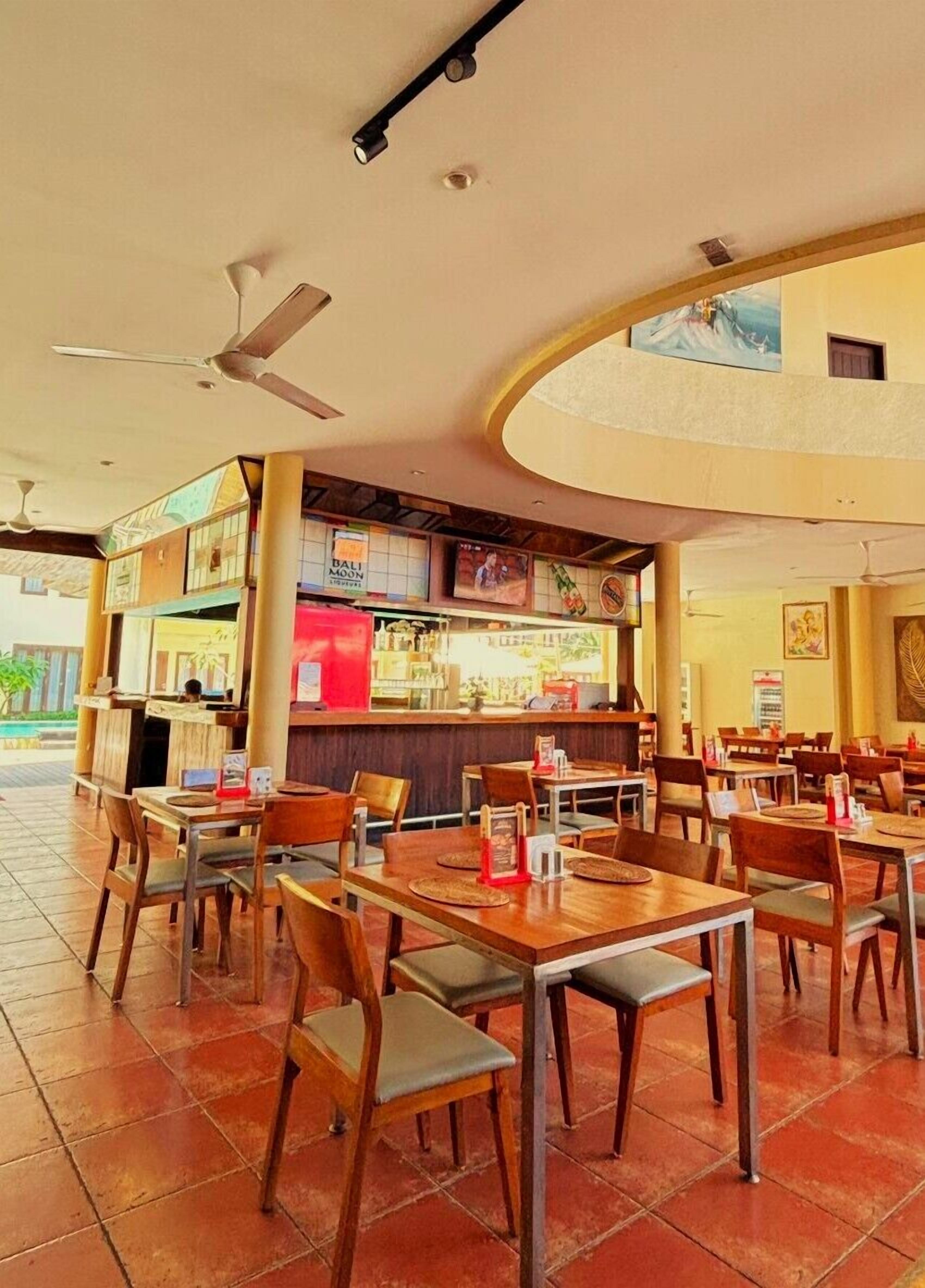 Um restaurante com mesas e cadeiras de madeira, um balcão com uma placa vermelha e um ventilador de teto.