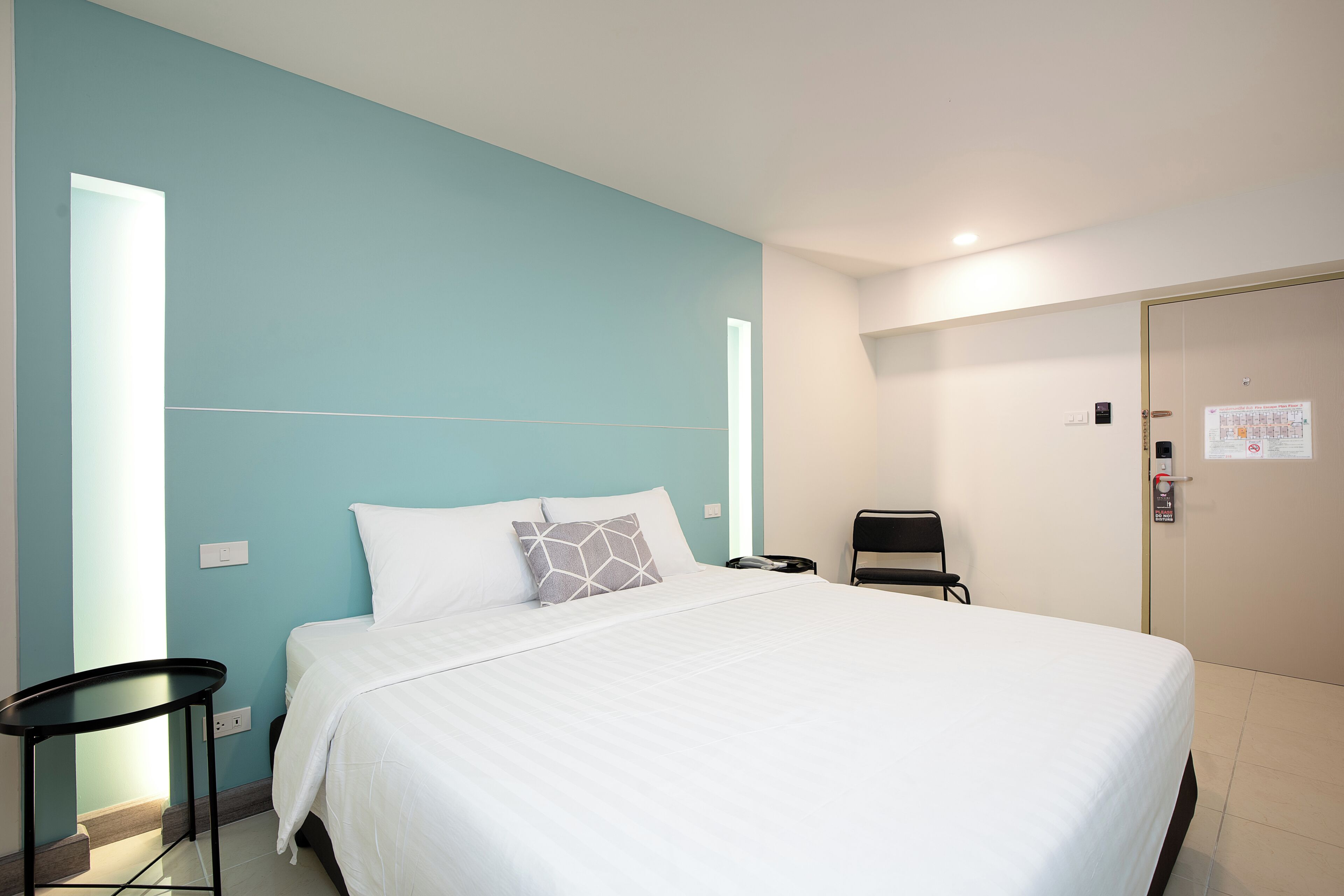 Quarto de hotel com cama, uma mesinha, uma cadeira e uma porta com um aviso.