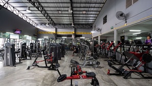 Uma academia com diversos equipamentos de exercício, teto alto e sinalização de saída visível.