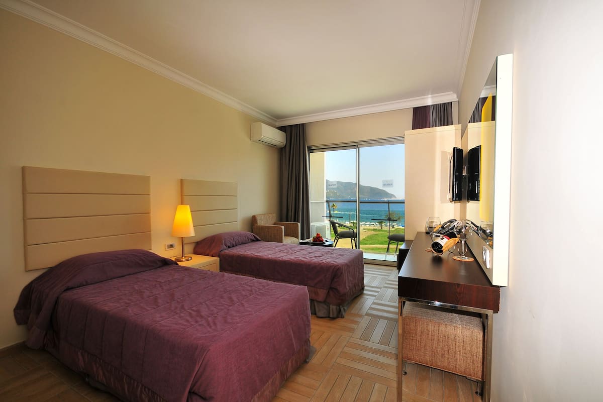 Quarto de hotel com duas camas, uma escrivaninha, uma cadeira e vista para o mar.