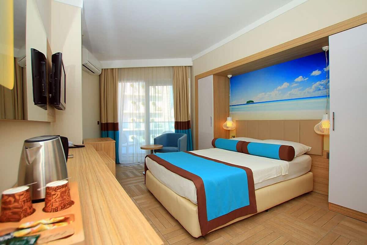 Quarto de hotel com uma cama, uma escrivaninha com cafeteira, uma cadeira, uma televisão e um quadro de uma praia.