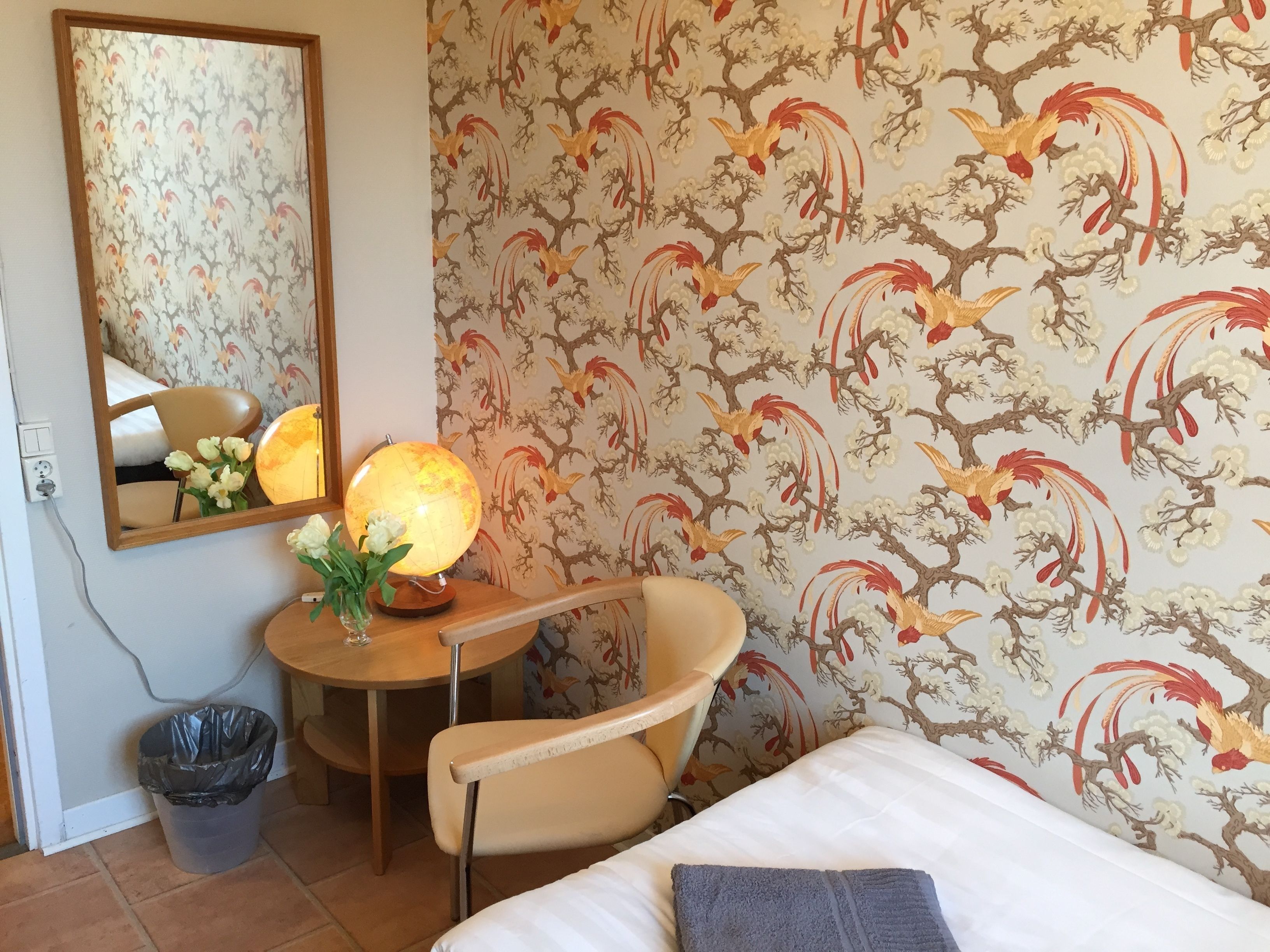Quarto de hotel com cama, escrivaninha com luminária globo, cadeira e lixeira.
