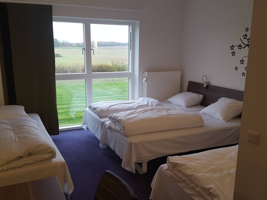 Quarto de hotel com duas camas de solteiro, uma janela grande com vista para um campo verde e uma luminária fixada na parede.