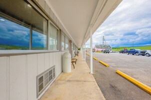 Área externa coberta com uma fileira de bancos, uma lixeira e vista para um estacionamento com veículos e um edifício ao fundo.