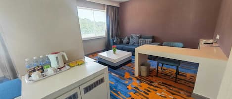 Quarto de hotel com sofá, mesa de centro, pequena mesa de jantar, cozinha compacta e uma janela grande.