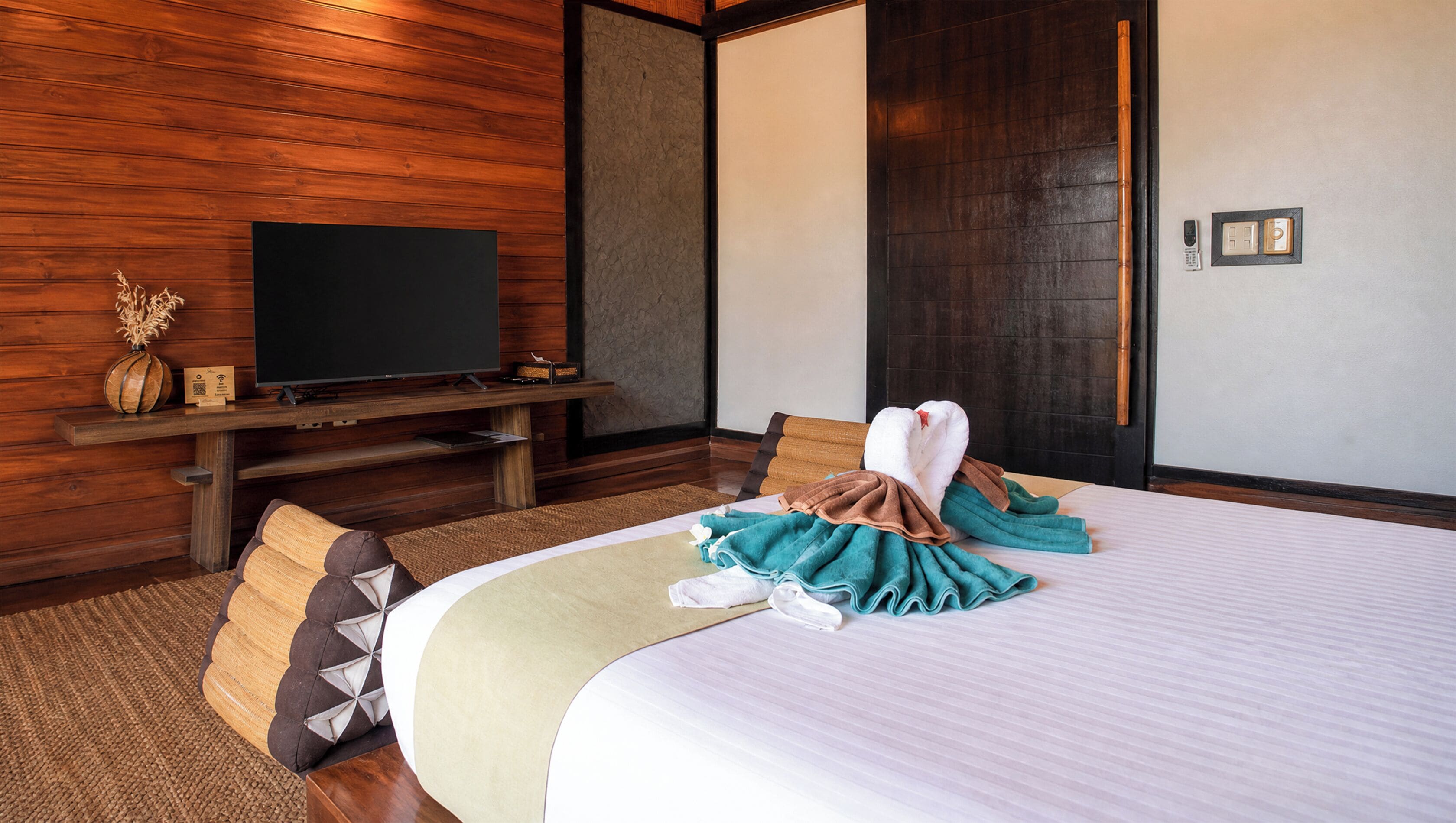 Quarto de hotel com cama, televisão de tela plana, parede de madeira, porta escura e interruptor de luz fixado na parede.