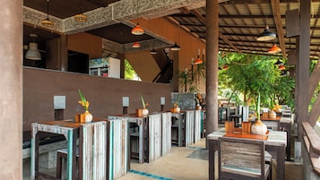 Restaurante com mesas e cadeiras de madeira, luminárias pendentes e plantas em vasos.