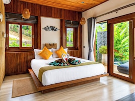 Quarto com estrutura de madeira, cama, duas almofadas, uma obra de arte na parede e vista para o verde pela janela.