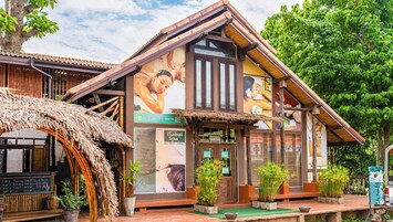 Edifício tradicional com telhado de palha, um cartaz grande anunciando serviços de spa e uma placa indicando comodidades como massagens e aromaterapia.